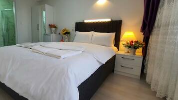 Cama bem arrumada com roupa de cama branca, abajur e uma mesinha de cabeceira com um vaso de flores.