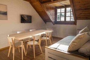 Quarto com mesa de jantar e cadeiras de madeira, cama com almofadas, TV fixada na parede e janela com vista para árvores.