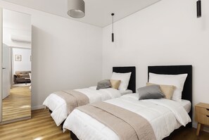Quarto moderno com duas camas, piso de madeira e porta que dá para outro cômodo.