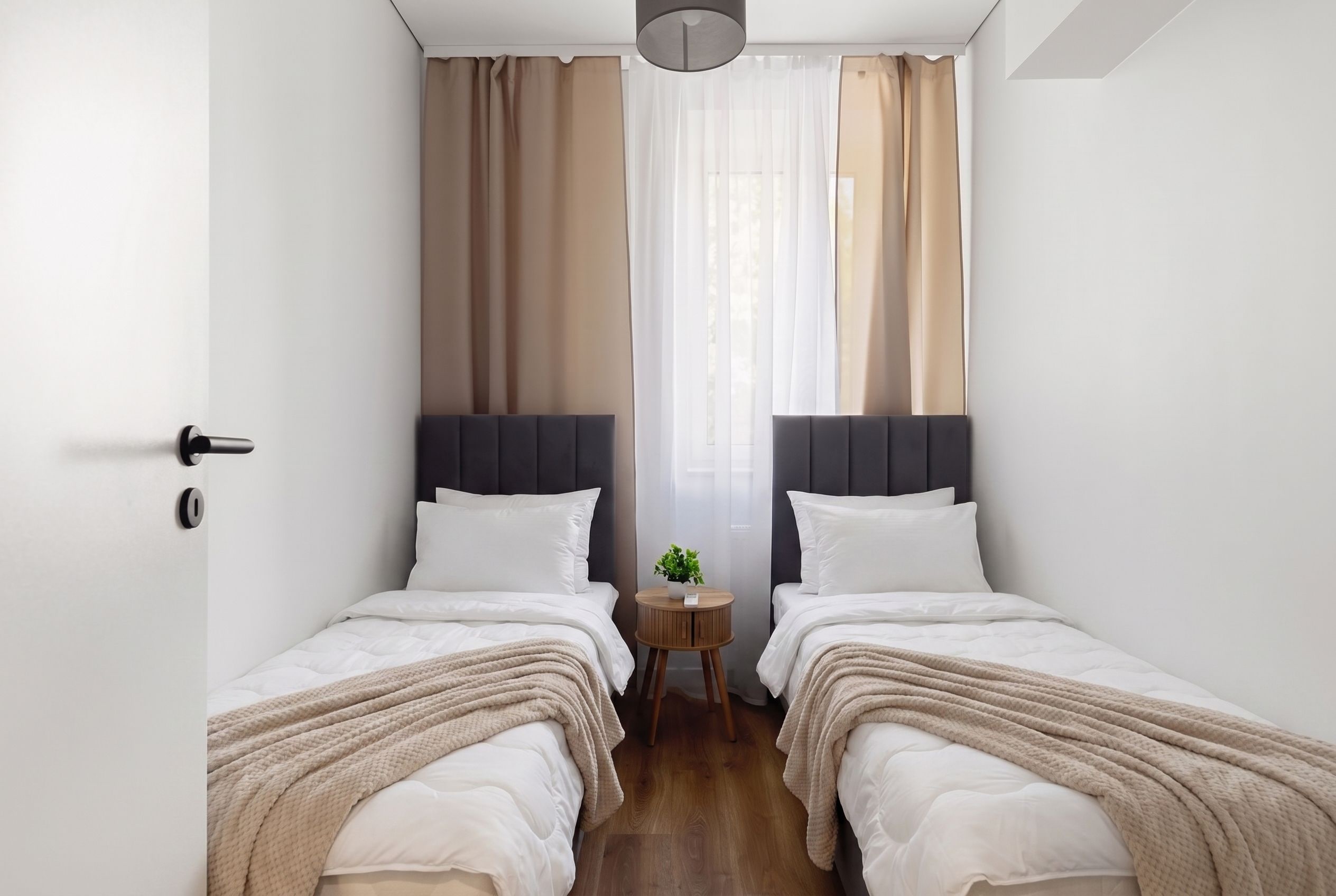 Quarto de hotel com duas camas, uma mesinha com uma planta e vista através de cortinas finas.