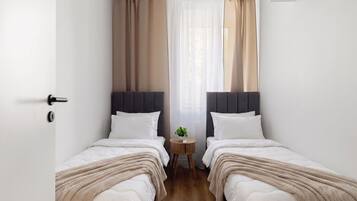 Quarto de hotel com duas camas, uma mesinha com uma planta e vista através de cortinas finas.