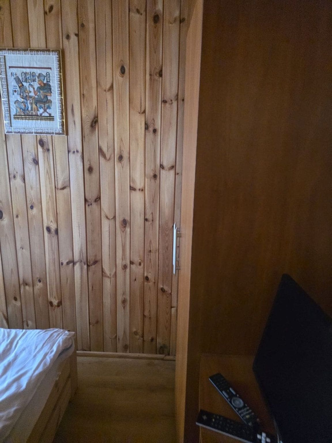 Quarto com painéis de madeira, uma cama, uma porta e um quadro na parede.