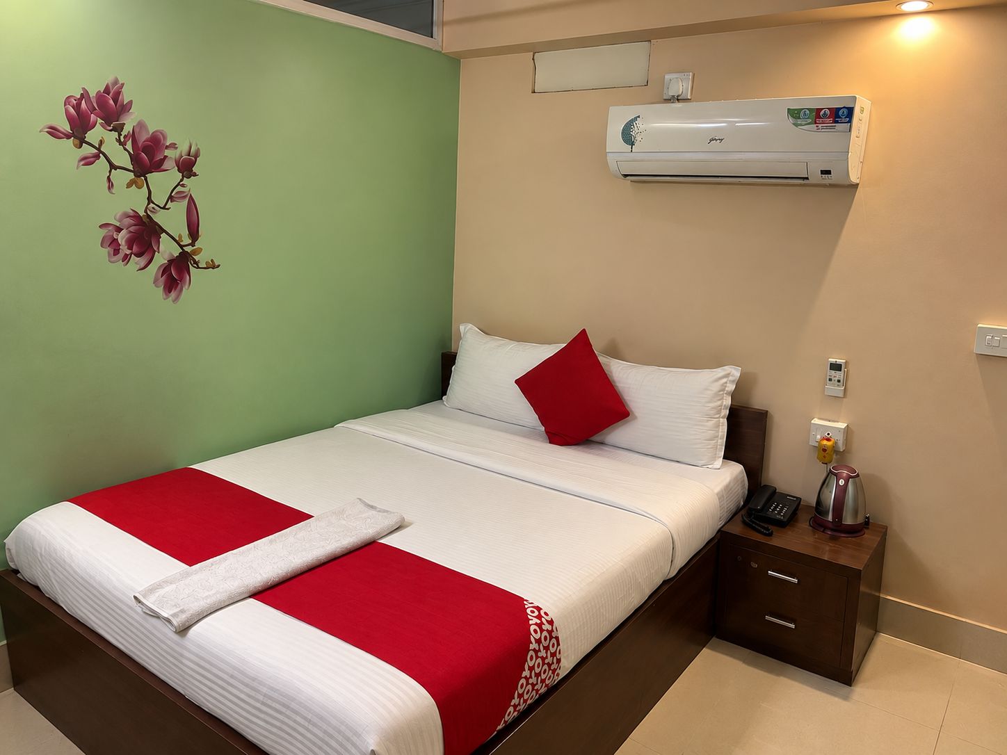 Quarto de hotel com cama, colcha vermelha e branca, mesa de cabeceira com telefone e chaleira, e unidade de ar condicionado na parede.
