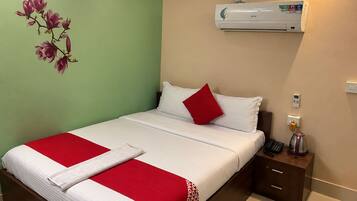 Quarto de hotel com cama, colcha vermelha e branca, mesa de cabeceira com telefone e chaleira, e unidade de ar condicionado na parede.