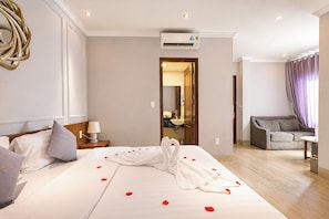 Quarto de hotel com cama grande, mesa de cabeceira com luminária, sofá e banheiro visível através de uma porta aberta.