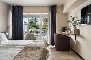 Quarto de hotel com cama, escrivaninha, cadeira, planta em vaso e janela com cortinas.