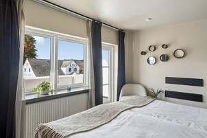 Quarto com uma janela grande, uma cama, um radiador e vista para uma casa lá fora.