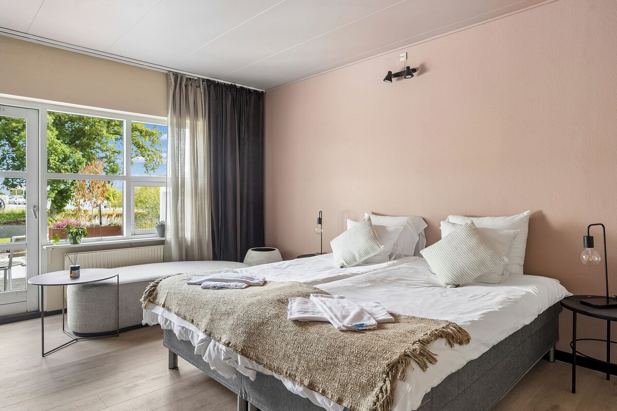 Quarto de hotel moderno com uma cama grande, mesas de cabeceira, uma janela com cortinas e vista para o verde lá fora.