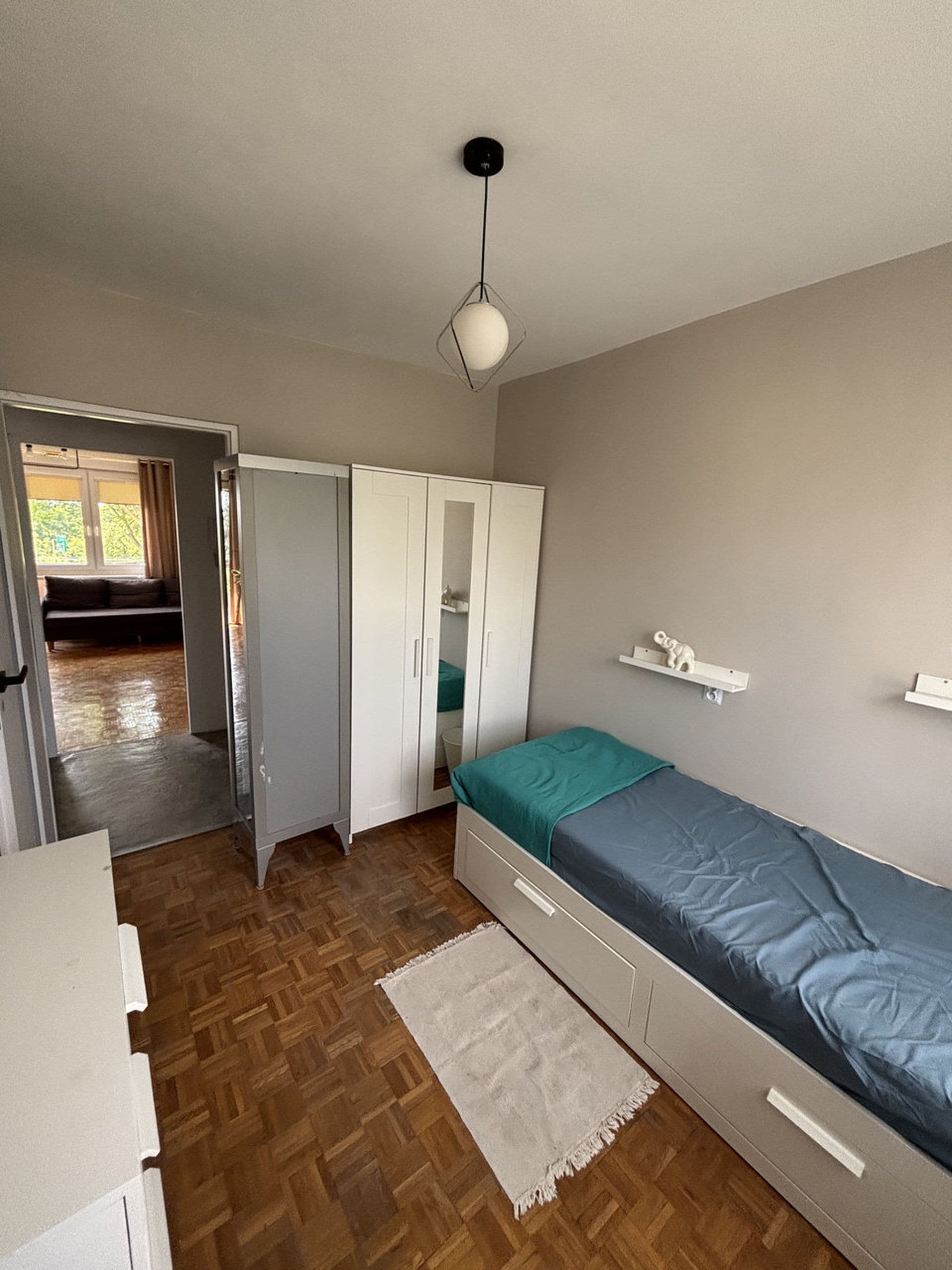 Quarto com cama, guarda-roupa, espelho, prateleira com uma estátua e porta que dá para outro cômodo.