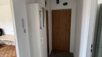 Corredor estreito com uma porta de madeira no fim, uma geladeira branca e uma pequena placa verde na parede.