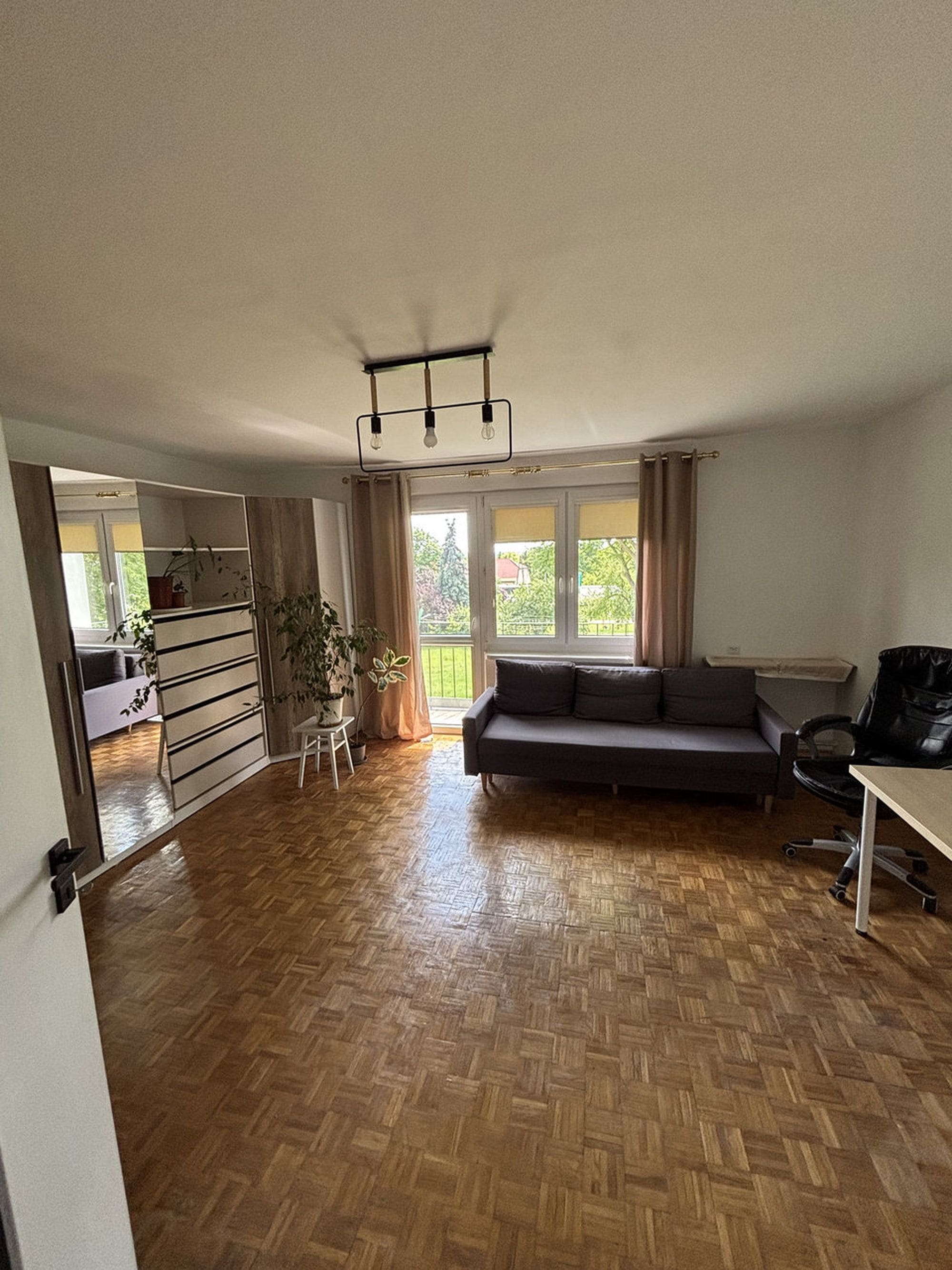 Ampla sala de estar com piso de madeira em padrão herringbone, um sofá escuro, uma mesa de jantar e um vaso com planta.