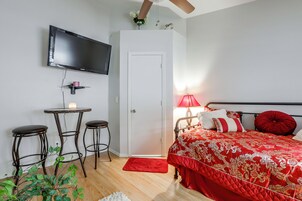 Quarto com cama, televisão presa na parede, escrivaninha com dois banquinhos, porta e tapete vermelho.
