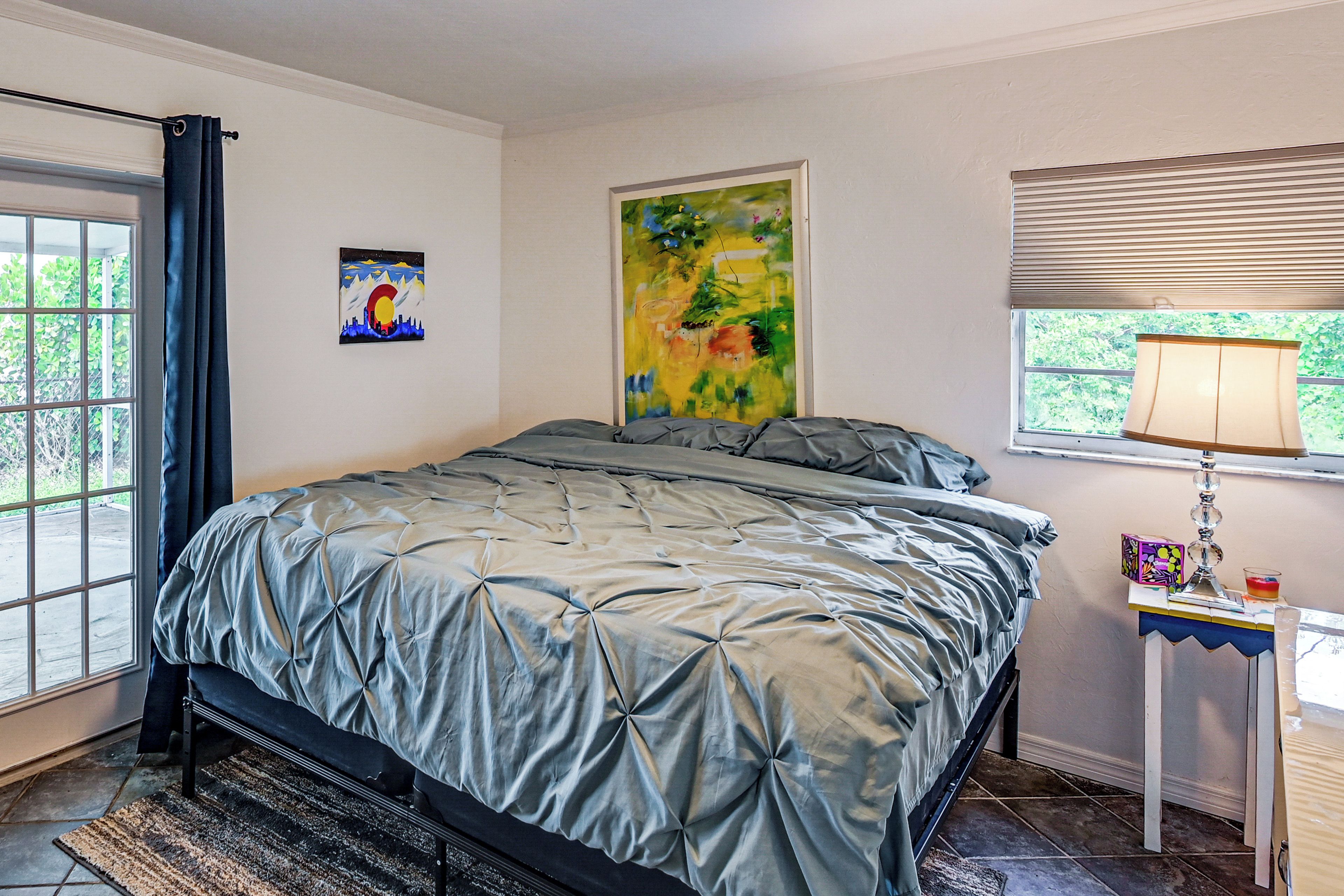 Quarto com cama, janela com persianas, um quadro na parede e abajur em uma mesinha de cabeceira.