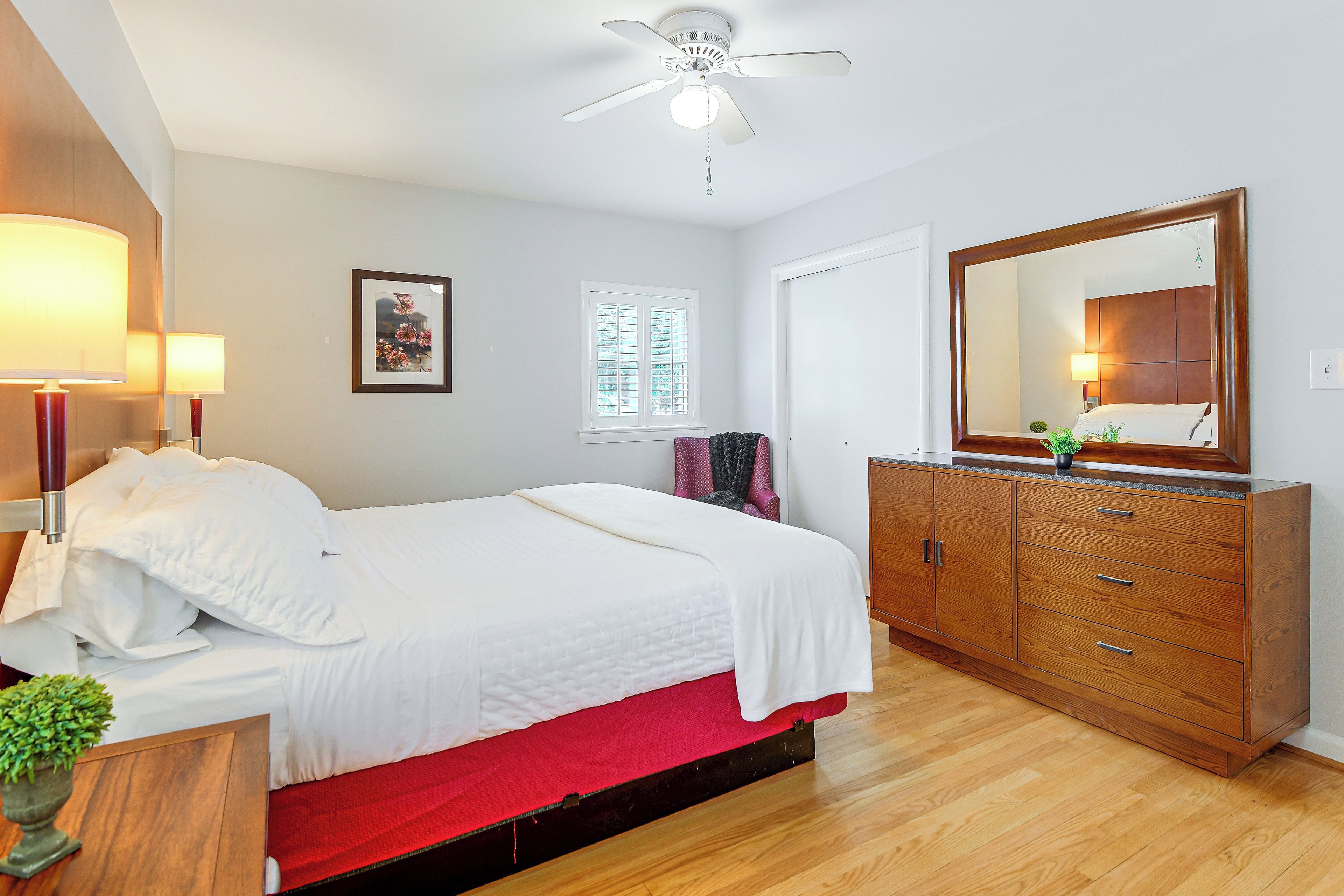 Quarto de hotel com cama, penteadeira, espelho, ventilador de teto e janela com persianas.