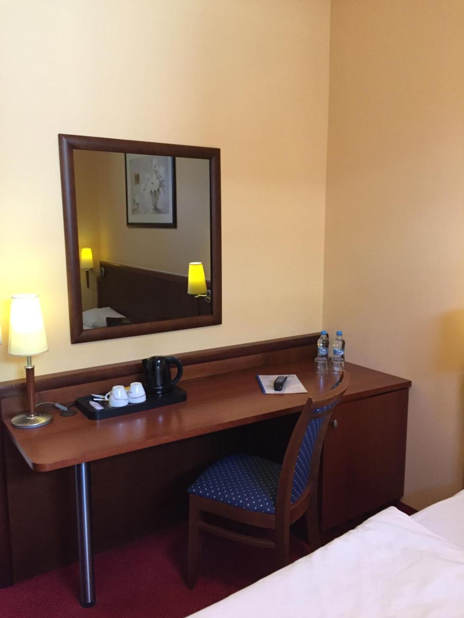 Quarto de hotel com escrivaninha de madeira, uma cadeira, cama, espelho, abajur e um quadro na parede.