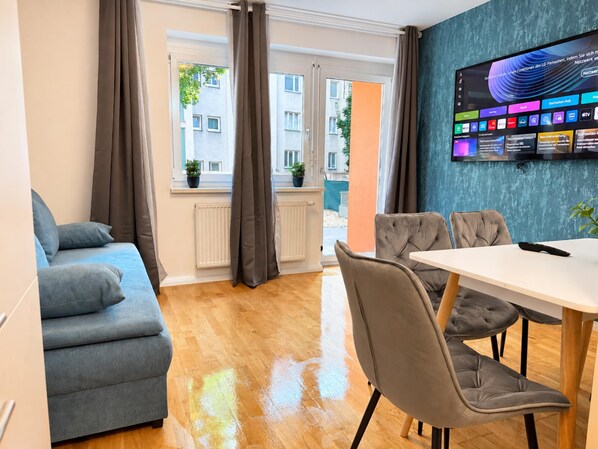 Uma sala de estar moderna com um sofá cinza, uma mesa de jantar com cadeiras, uma TV fixada na parede e vista para o exterior.