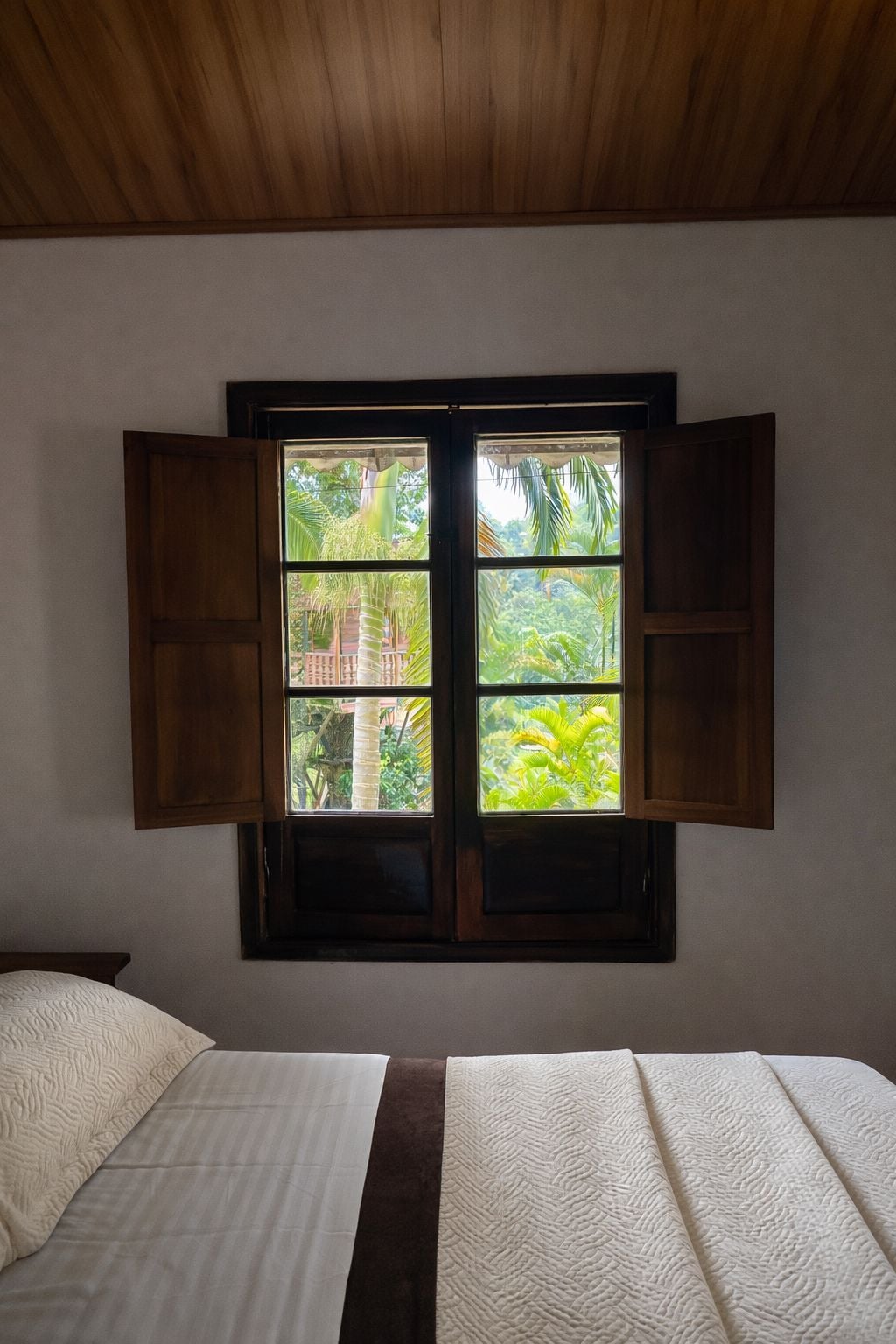 Quarto com cama, janela com persianas abertas e vista para o verde lá fora.