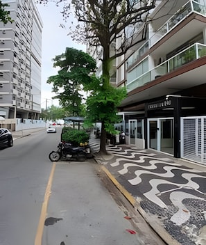 Uma rua com calçada arborizada, uma bicicleta estacionada e um prédio com loja.
