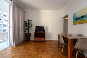 Uma sala de estar moderna com piso de madeira em padrão herringbone, uma janela grande com cortinas, um vaso de planta, uma TV de tela plana em um suporte de madeira, uma mesa de jantar com cadeiras e um quadro na parede.
