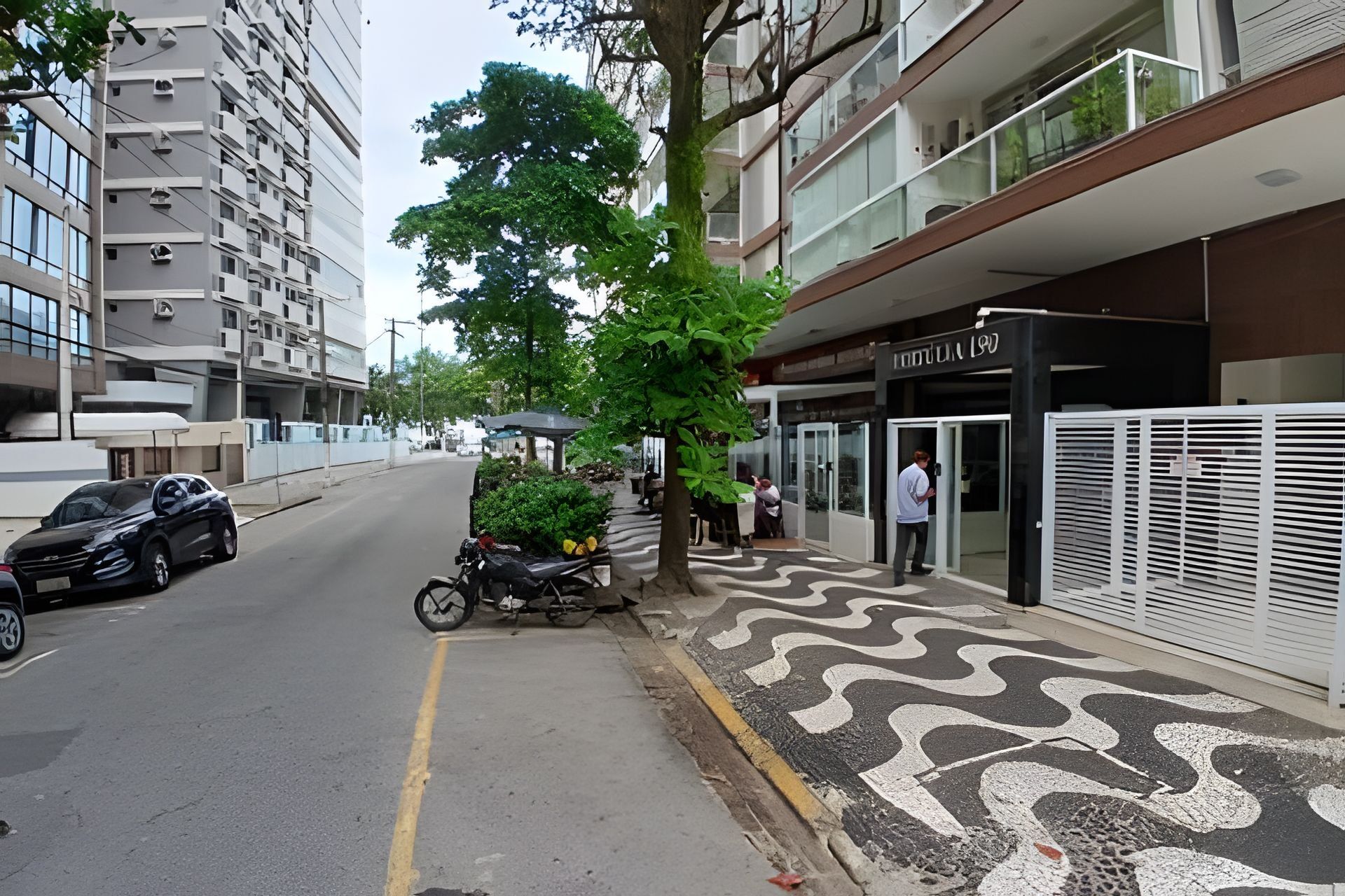 Rua com um carro estacionado e uma motocicleta, calçada arborizada e um prédio com fachada comercial.
