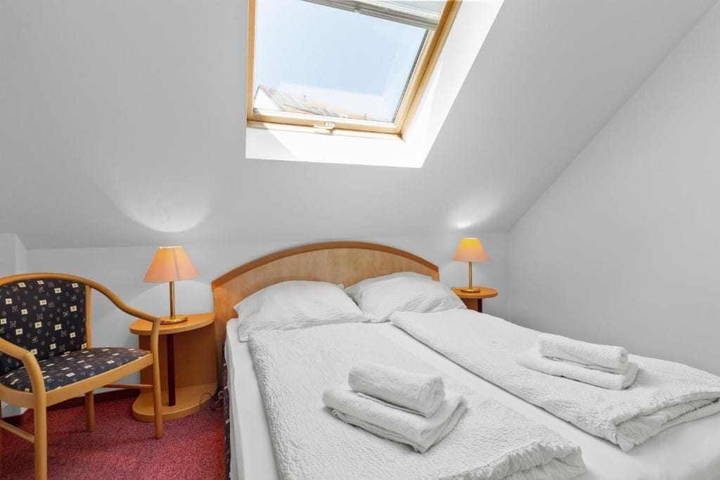 Quarto de hotel com uma cama de solteiro, duas luminárias de cabeceira, uma cadeira e um claraboia.
