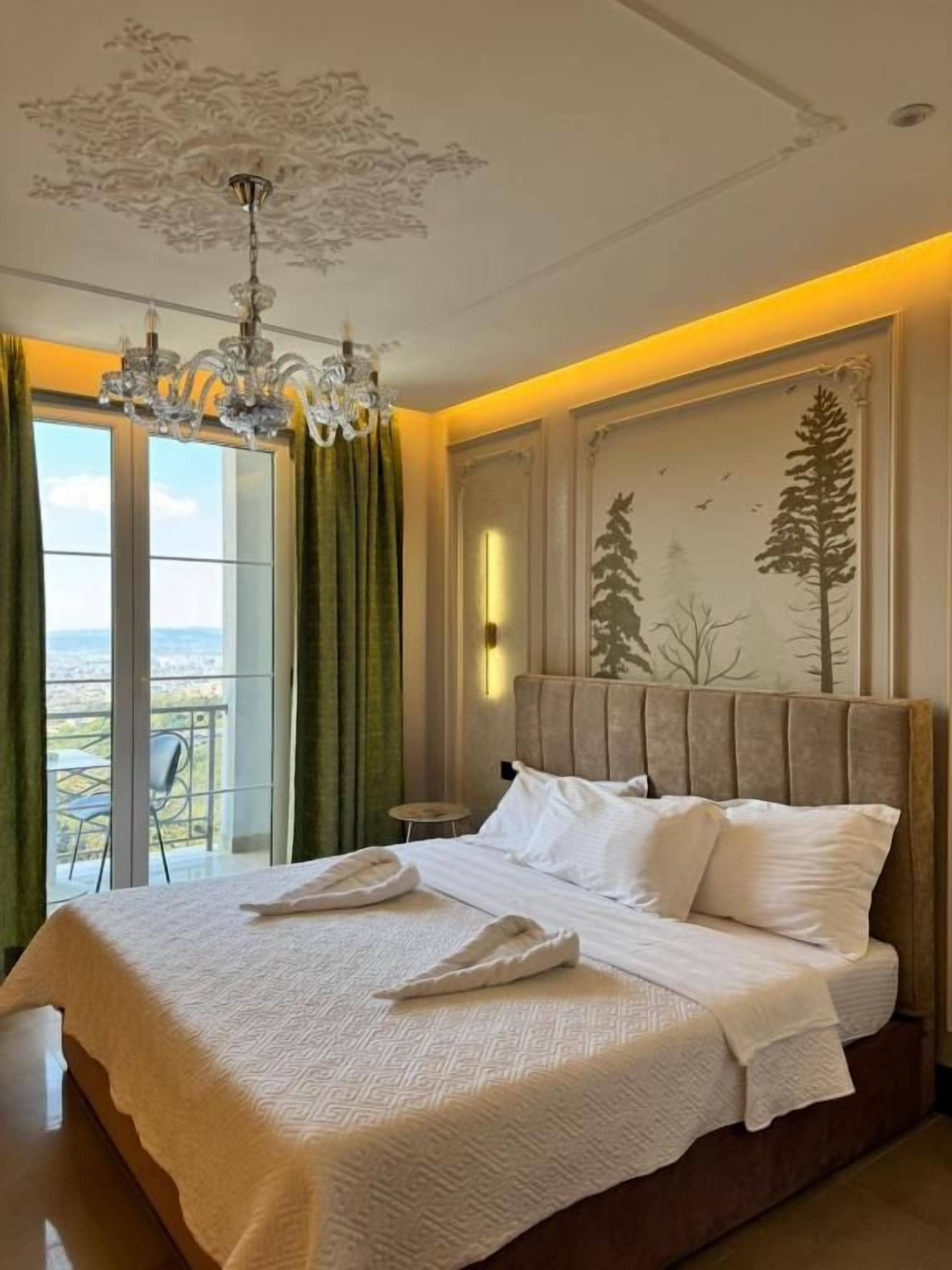 Quarto de hotel com cama grande, um lustre, vista através de uma porta de vidro e parede com desenho de árvore.