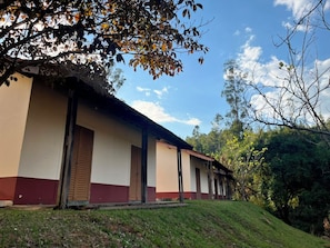 Um edifício com vários quartos, cada um com porta e janela. O edifício tem telhado plano e é cercado por vegetação.
