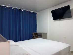 Quarto com cama, escrivaninha, cadeira, televisão fixada na parede e janela com cortinas azuis.