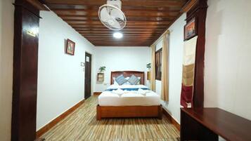 Quarto de hotel com teto de madeira, cama com estrutura de madeira, mesa de cabeceira e ventoinha fixada na parede.