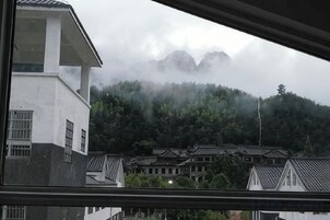 Um edifício tradicional com fachada branca e telhado inclinado, rodeado por vegetação e montanhas ao fundo.