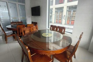 Uma sala de jantar com uma mesa de vidro redonda cercada por cadeiras de madeira, uma televisão na parede e uma janela com vista para a rua.