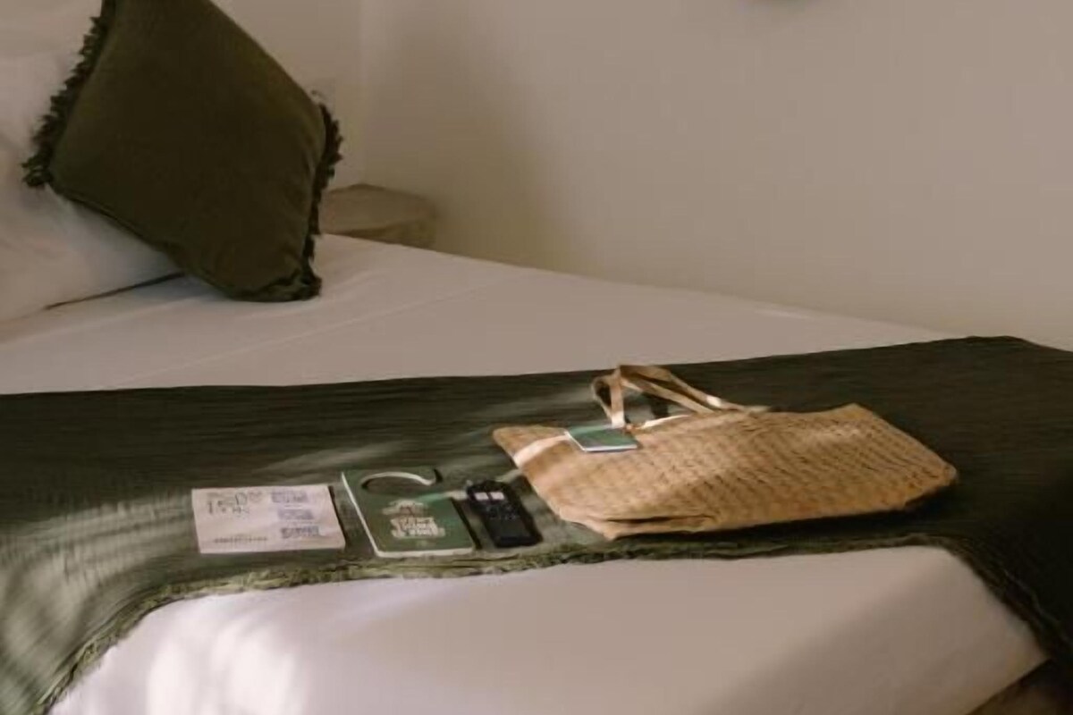 Uma cama com um cobertor verde, uma cesta de palha e um luminário fixado na parede.