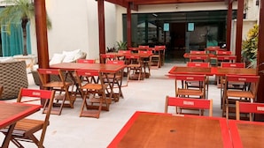 Um café ao ar livre com várias mesas e cadeiras de madeira, algumas com almofadas vermelhas.