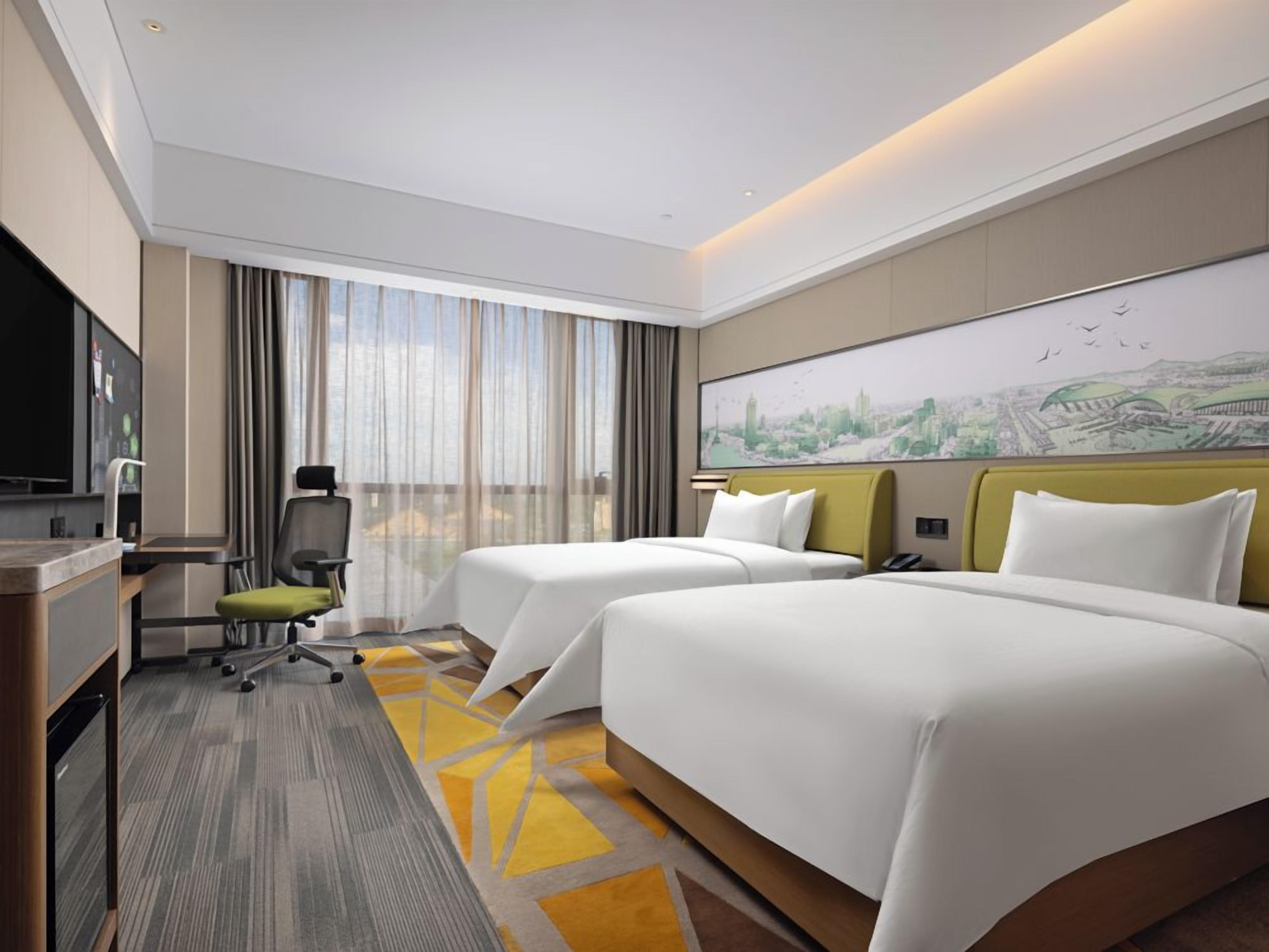 Quarto de hotel com duas camas, uma escrivaninha, uma cadeira, uma televisão e uma janela grande com cortinas.