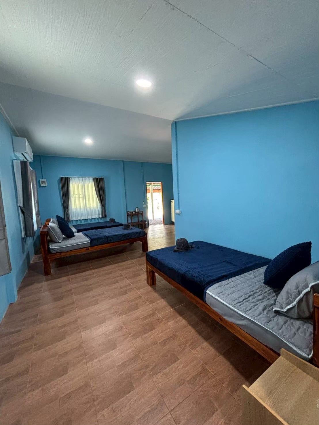 Quarto com duas camas de madeira, janela com cortinas, ventoinha no teto e piso de madeira.