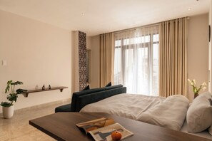 Quarto de hotel moderno com uma cama grande, um sofá escuro, uma mesa de centro de madeira e uma planta em vaso.