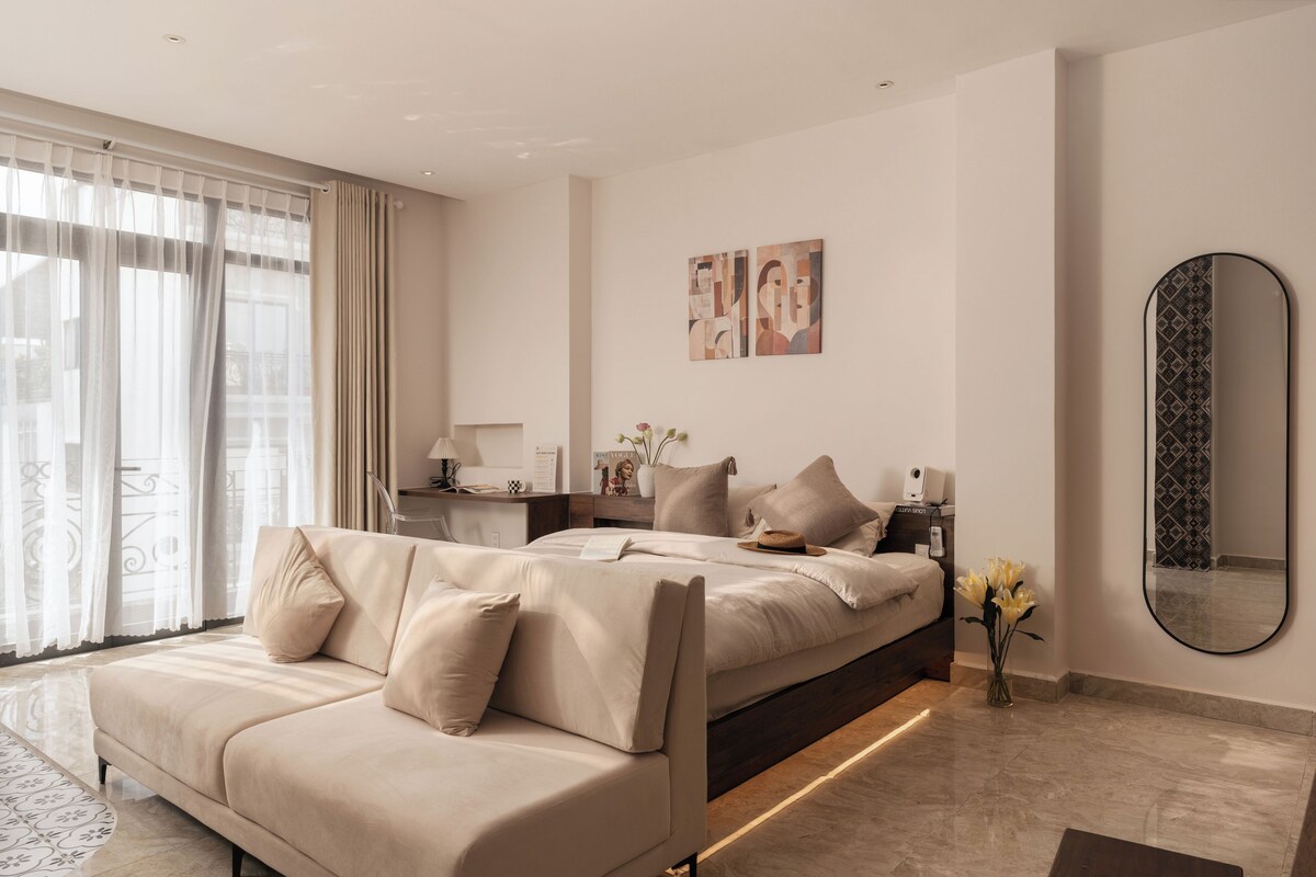 Quarto moderno com cama, sofá, mesa de cabeceira, espelho e janela com cortinas.