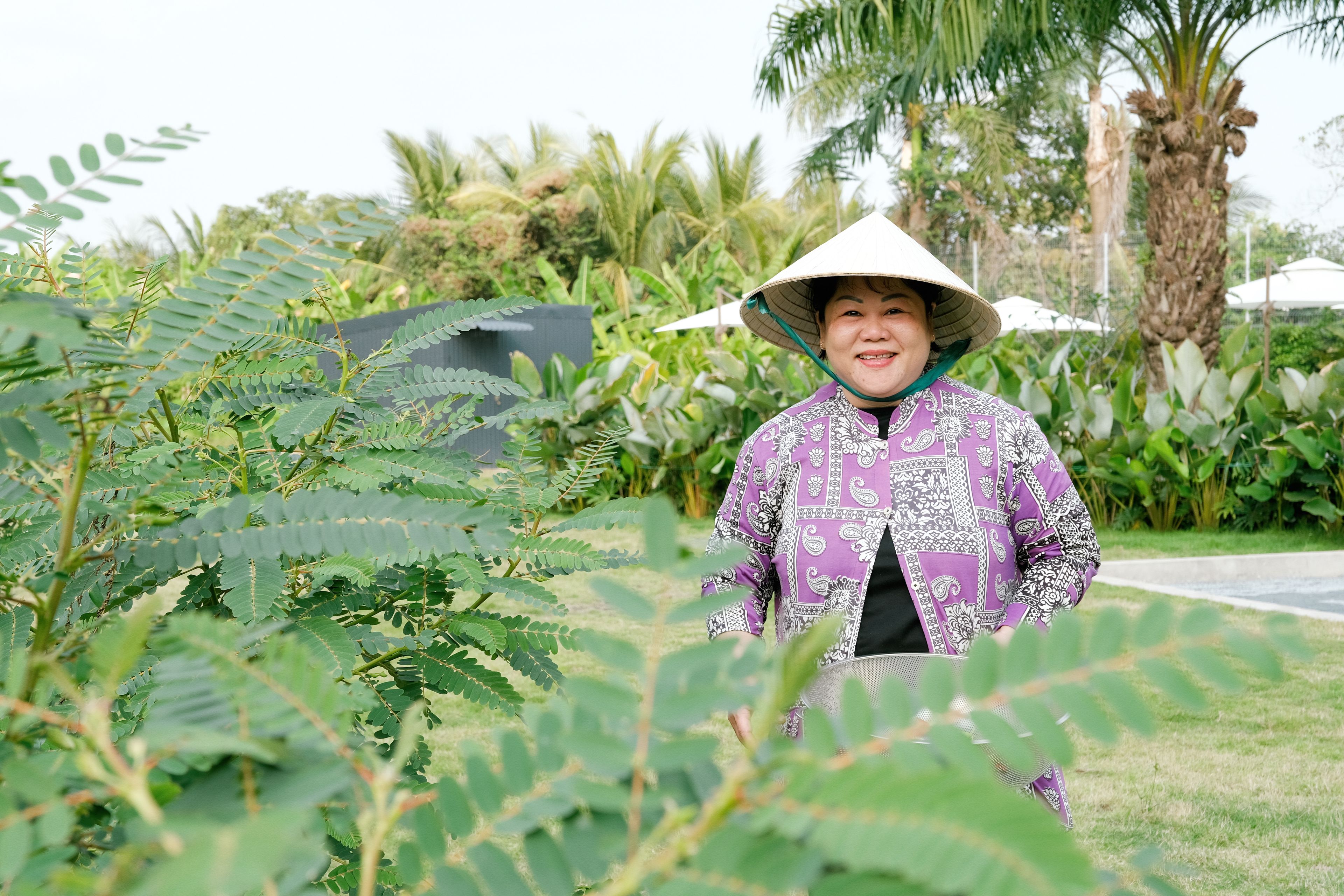 Uma pessoa vestindo um casaco estampado e um chapéu cônico, parada em um jardim com diversas plantas verdes e uma palmeira ao fundo.