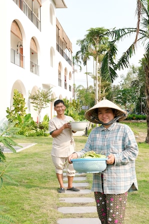 Pessoa segurando uma cesta de legumes, em uma área de jardim com um prédio ao fundo.