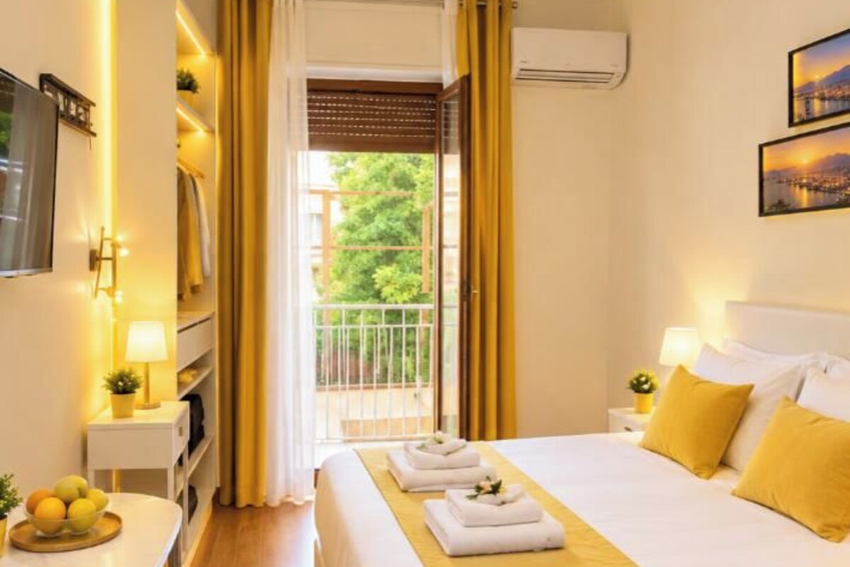 Quarto de hotel com cama, mesas de cabeceira, escrivaninha e varanda com cortinas.