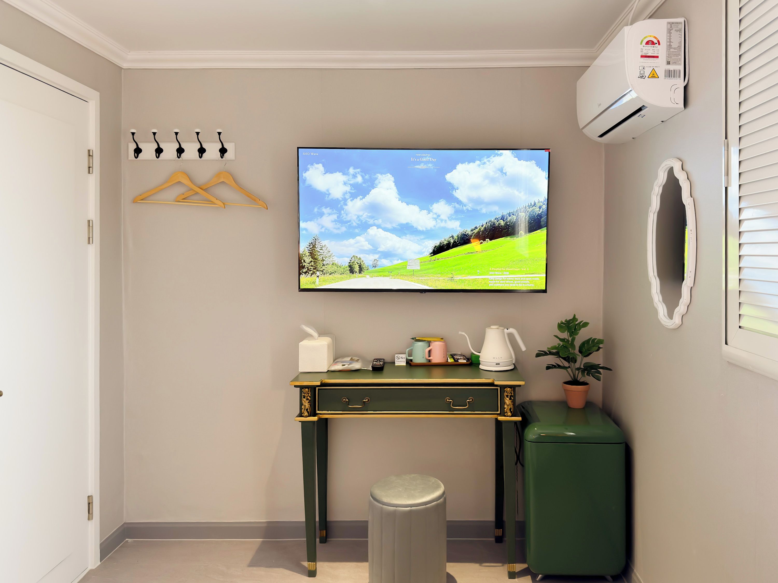Quarto com televisão na parede exibindo uma paisagem, uma mesa pequena com chaleira e xícara, lixeira e eletrodoméstico verde.