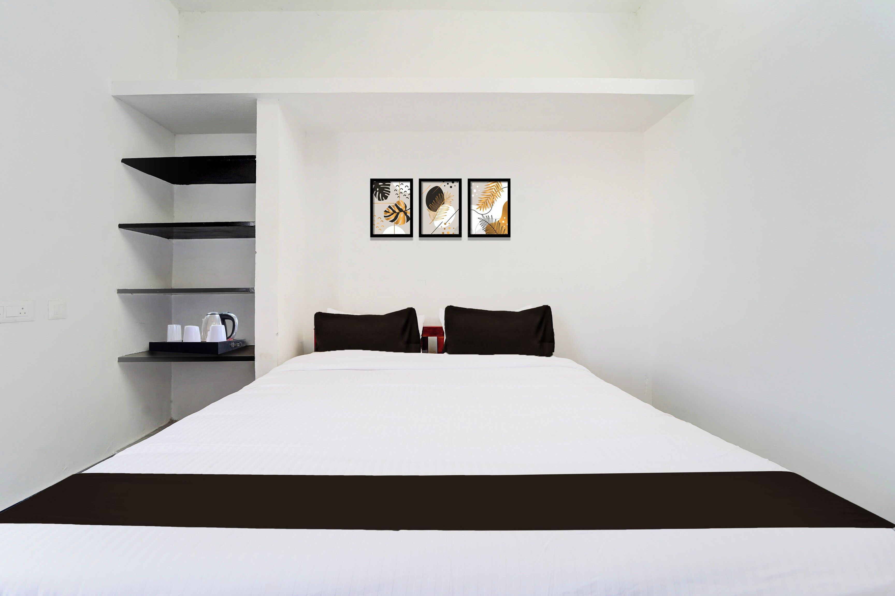Quarto de hotel com uma cama, uma prateleira com chávenas e uma chaleira, e três quadros na parede.