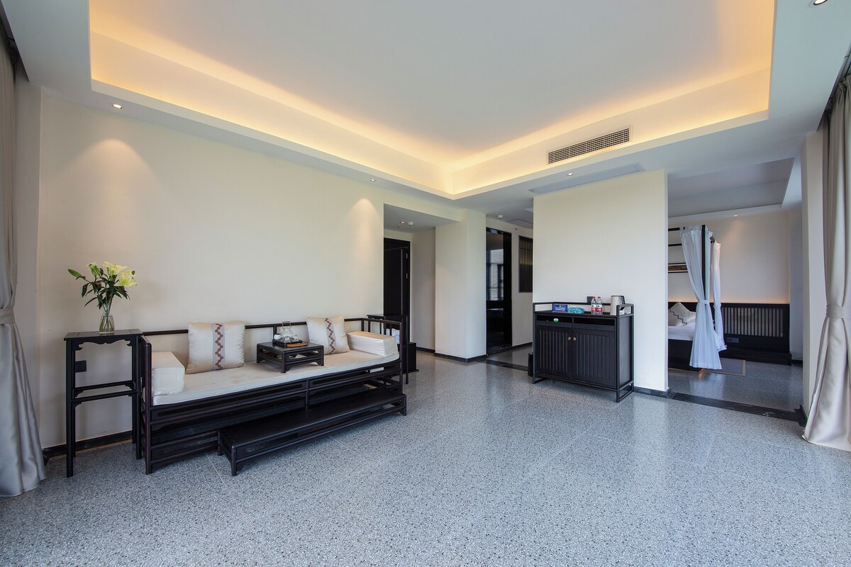 Um hall de hotel moderno com um sofá, uma mesa pequena e uma planta. Há uma porta que dá para um quarto com uma cama e um banheiro.