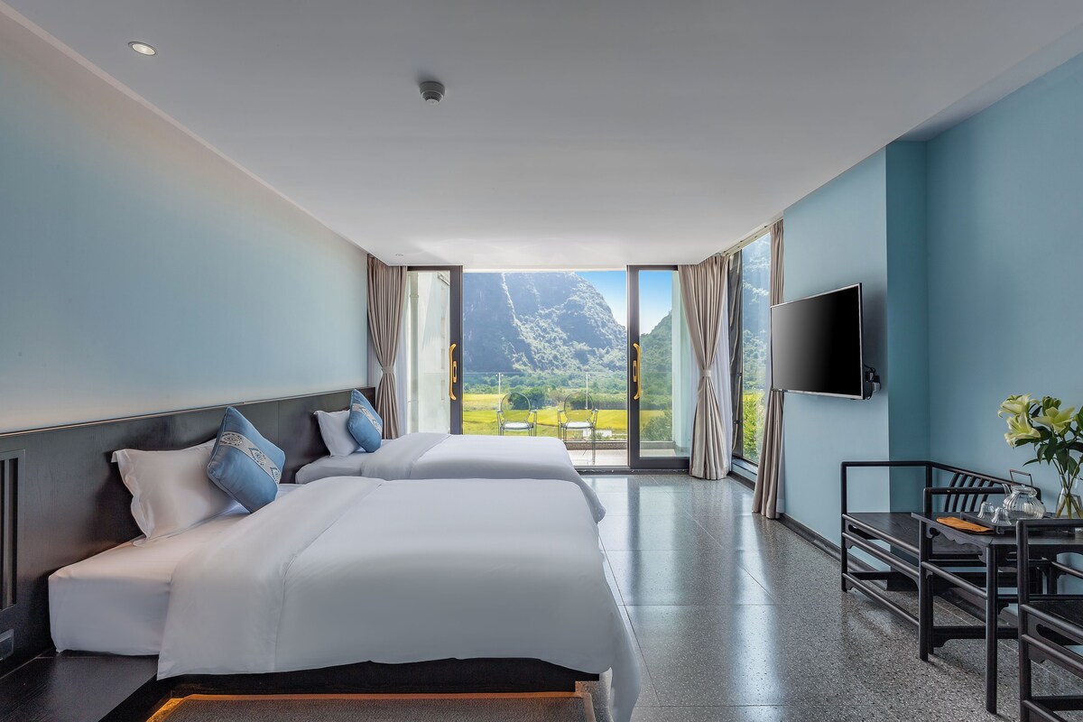 Quarto de hotel com duas camas, televisão, mesa com um vaso de flores e vista para montanhas.