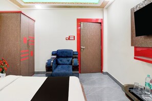 Quarto de hotel com uma cama, uma cadeira azul, uma TV e uma porta vermelha.