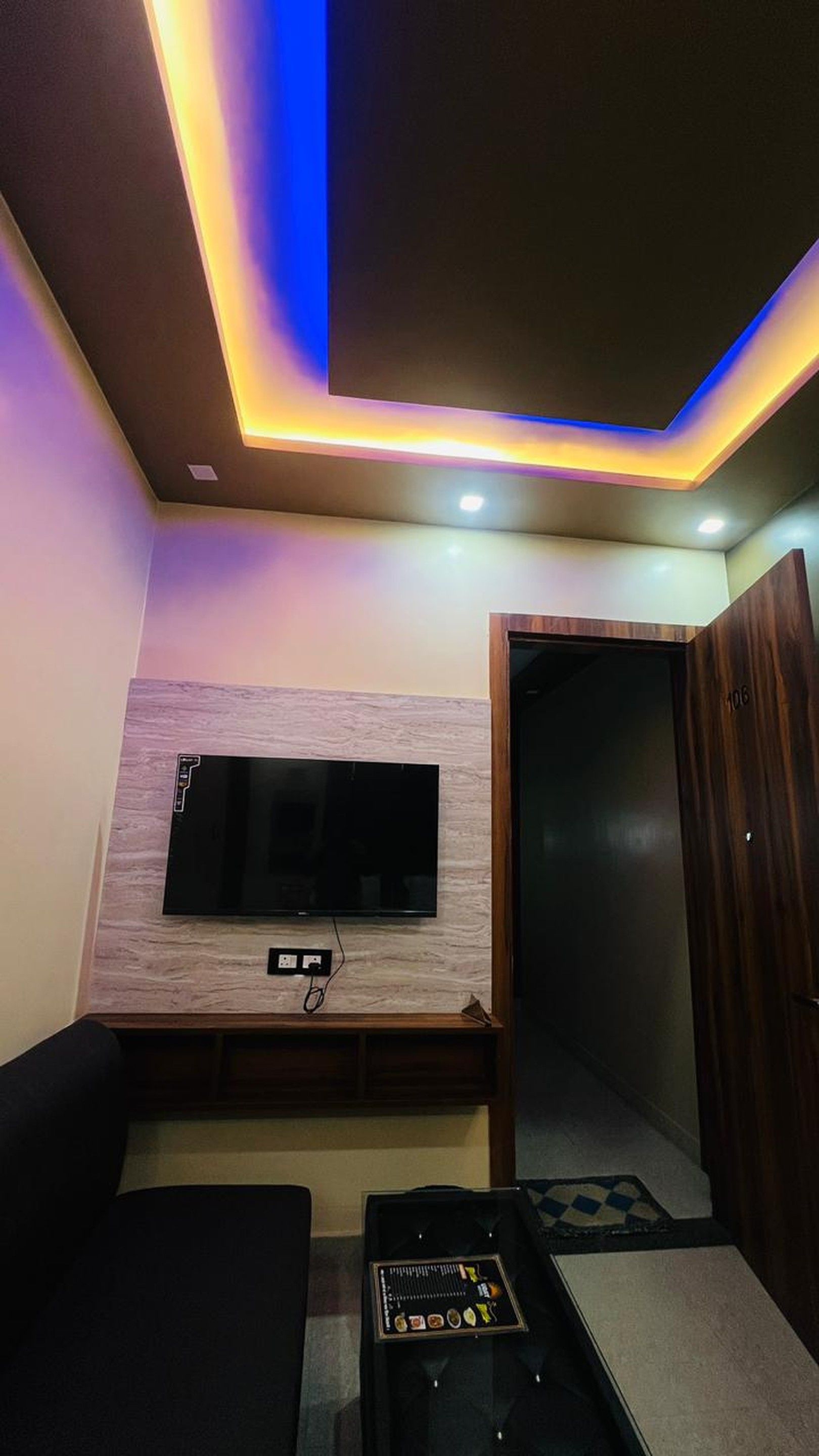 Uma sala de estar moderna com uma TV de tela plana fixada em uma parede texturizada, um sofá escuro, uma mesa de centro com dispositivos eletrônicos e uma porta de madeira.
