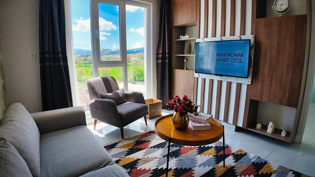 Sala de estar moderna com sofá, poltrona e mesa de centro com um vaso de flores. Uma TV de tela plana está instalada na parede, e há vista para o exterior por uma grande janela.