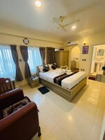 Quarto de hotel com cama, cadeira, escrivaninha, ventilador de teto e janela com cortinas.