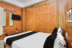 Quarto de hotel com parede de madeira, cama com roupa de cama branca e preta, televisão de tela plana na parede, escrivaninha de madeira com um vaso de flores e uma janela com cortinas.