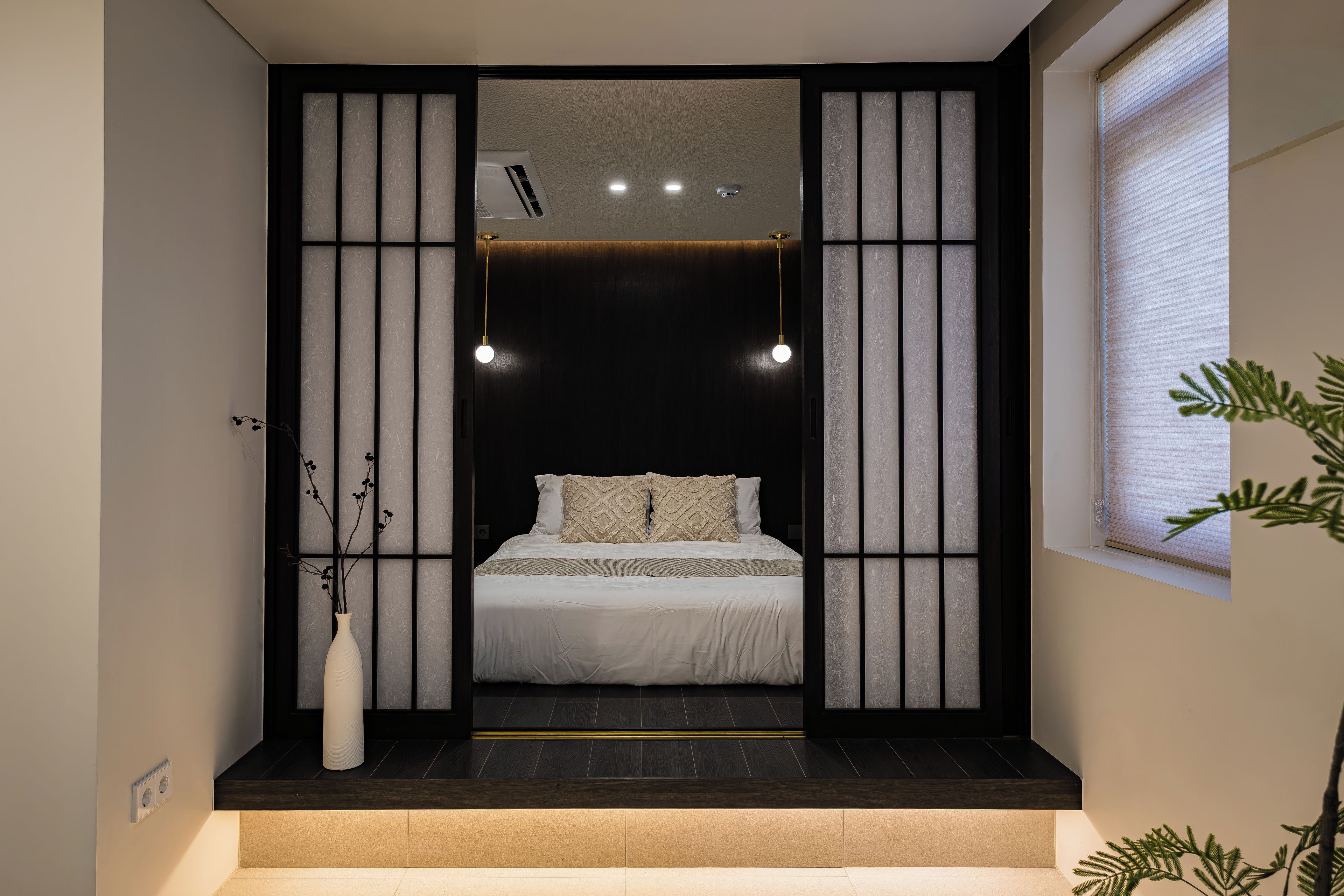 Quarto moderno com uma cama centralizada em um recanto escuro, com painéis de vidro fosco nas laterais, um vaso branco com galhos e uma janela com persianas.